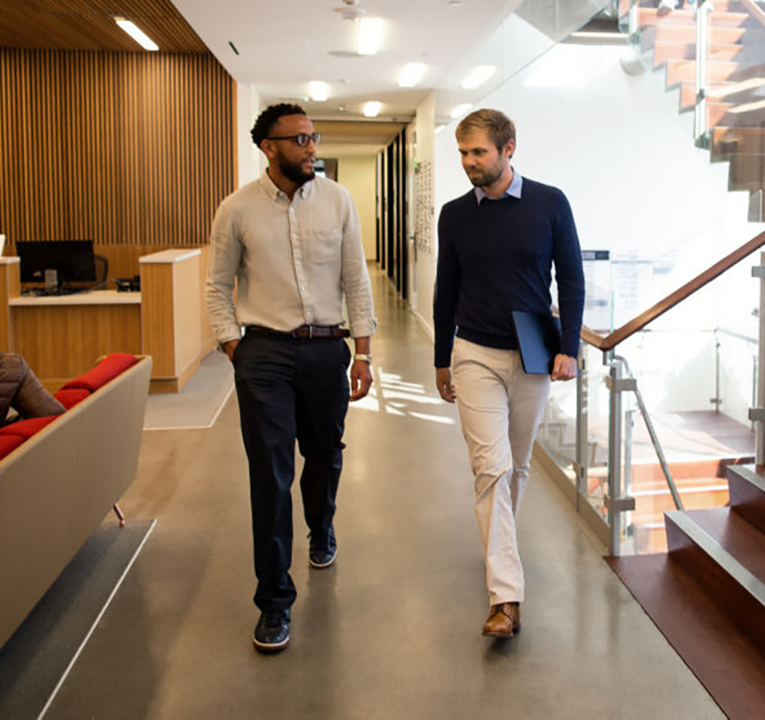 Two men taking as they walk down a corridor in an office building.